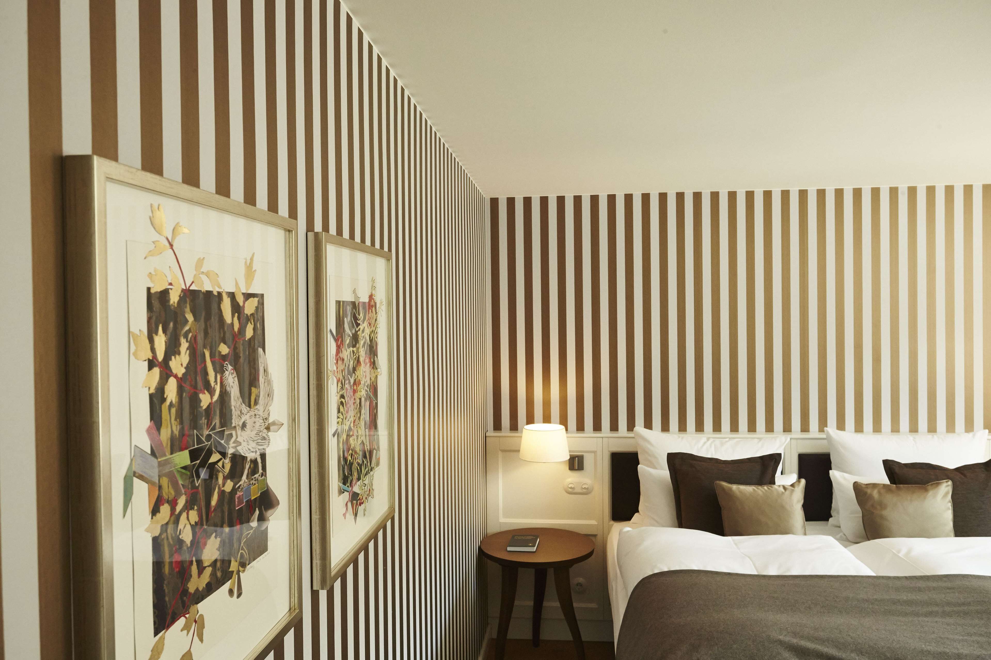 Quarto de hotel com papel de parede listrado, uma cama com várias almofadas, mesa de cabeceira com abajur e obras de arte emolduradas na parede.