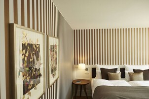 Quarto de hotel com papel de parede listrado, uma cama com várias almofadas, mesa de cabeceira com abajur e obras de arte emolduradas na parede.