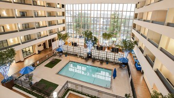 Um átrio de hotel com vários níveis, piscina central, plantas tropicais e áreas de estar.