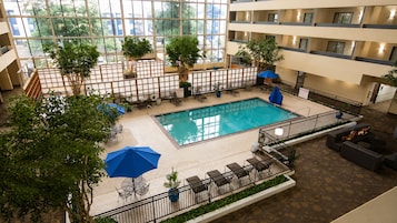 Uma piscina interna com piso de azulejos, cercada por espreguiçadeiras e guarda-sóis, e vista para o exterior através de grandes janelas.