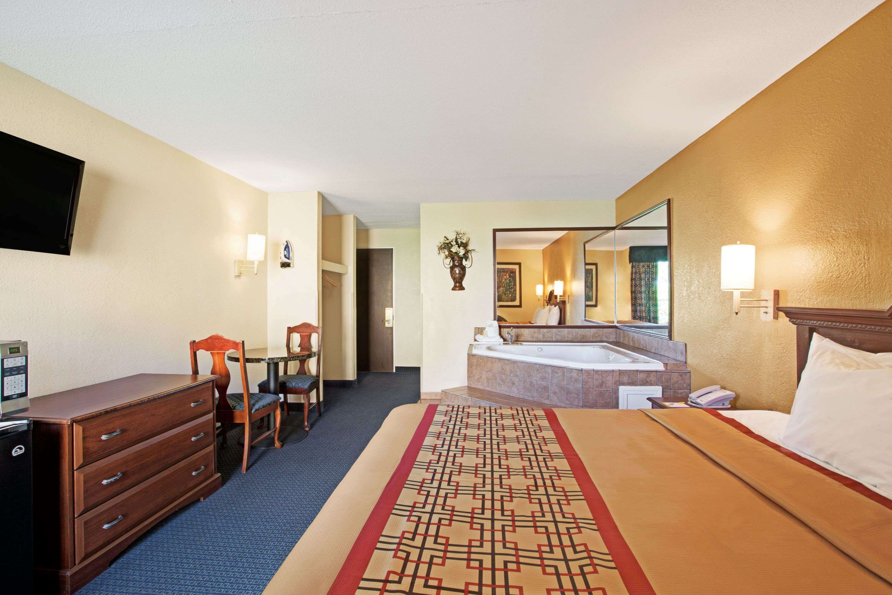 Quarto de hotel com cama, penteadeira, escrivaninha, cadeira, abajur, telefone, televisão, espelho e banheiro com banheira e pia.