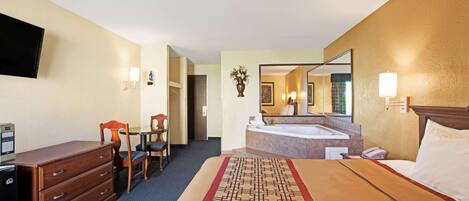 Quarto de hotel com cama, penteadeira, escrivaninha, cadeira, abajur, telefone, televisão, espelho e banheiro com banheira e pia.