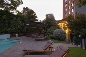 Uma área de piscina no terraço com espreguiçadeiras e guarda-sóis, cercada por vegetação e um edifício ao fundo.