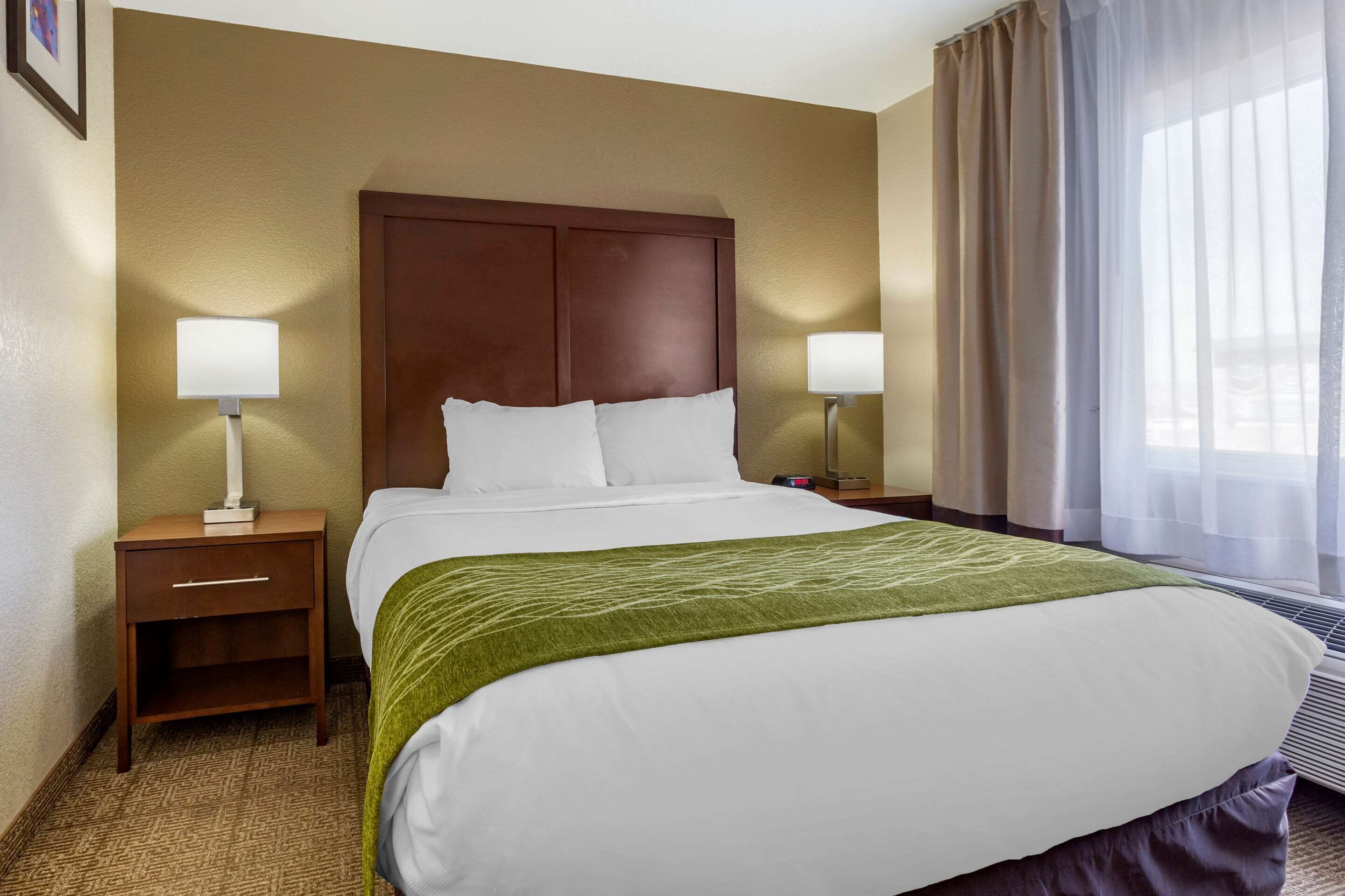 Quarto de hotel com uma cama grande, duas mesas de cabeceira com luminárias, uma cabeceira de madeira e uma janela com cortinas.