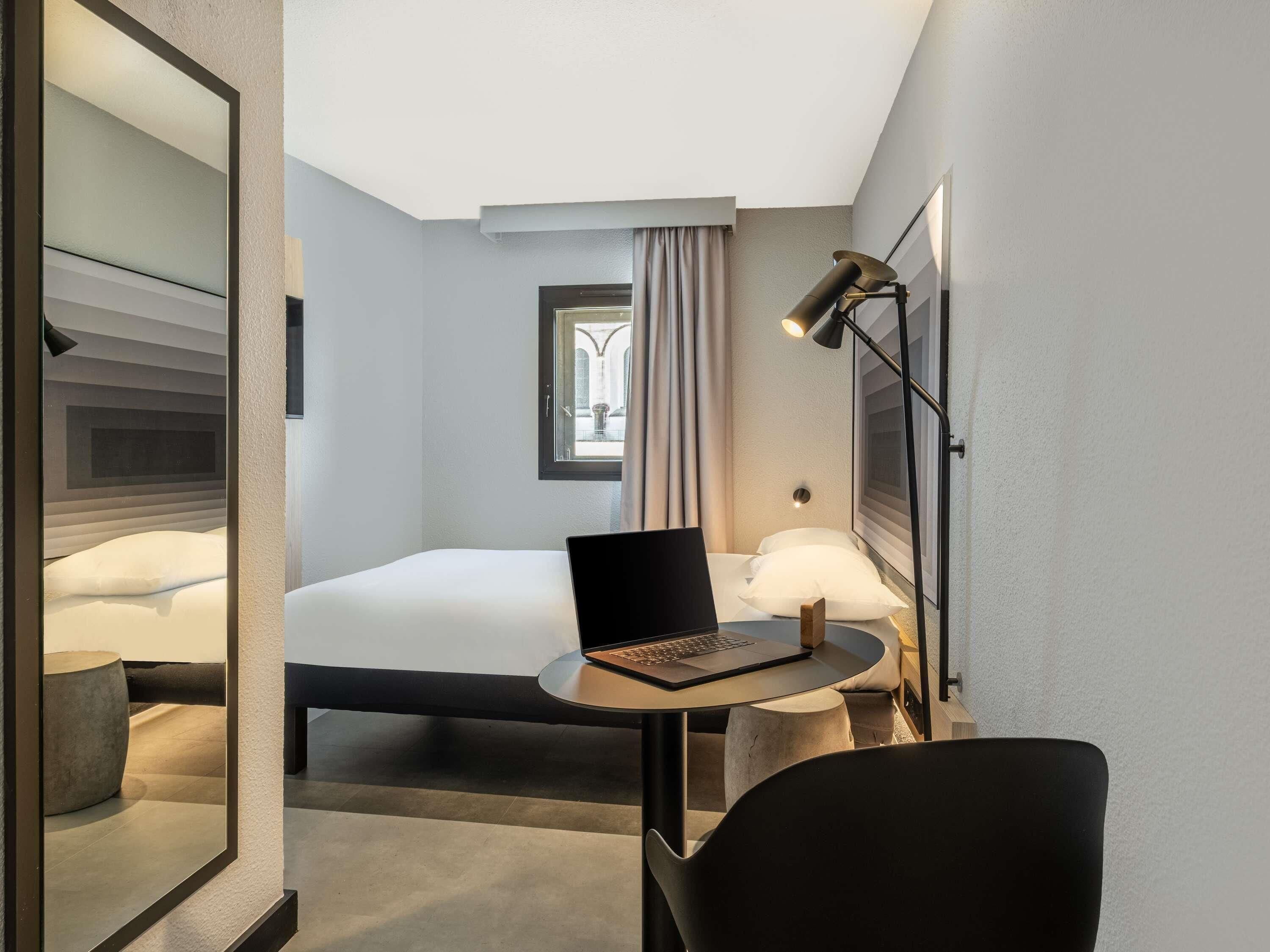 Quarto de hotel com cama, escrivaninha com um laptop, cadeira, abajur e janela com cortinas.