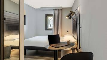 Quarto de hotel com cama, escrivaninha com um laptop, cadeira, abajur e janela com cortinas.