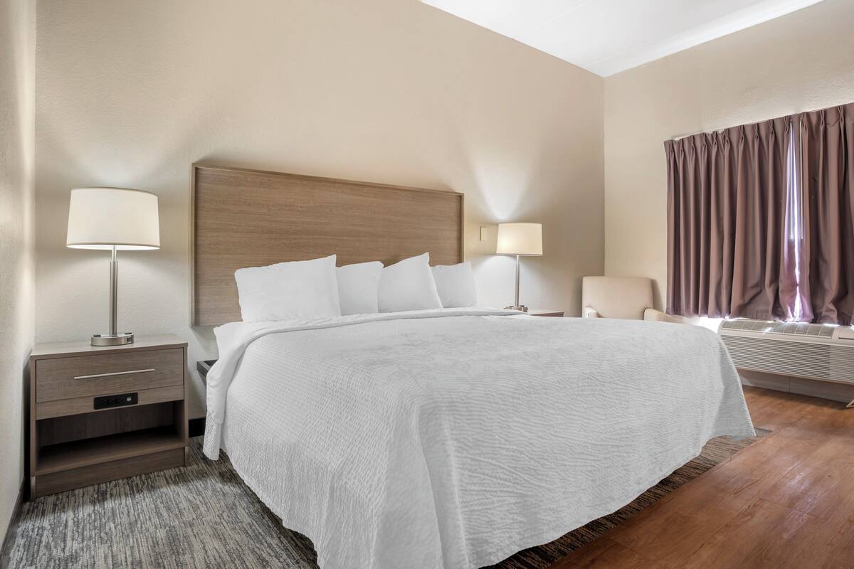Quarto de hotel com uma cama grande, duas mesas de cabeceira com luminárias, uma cadeira e uma janela com cortinas.