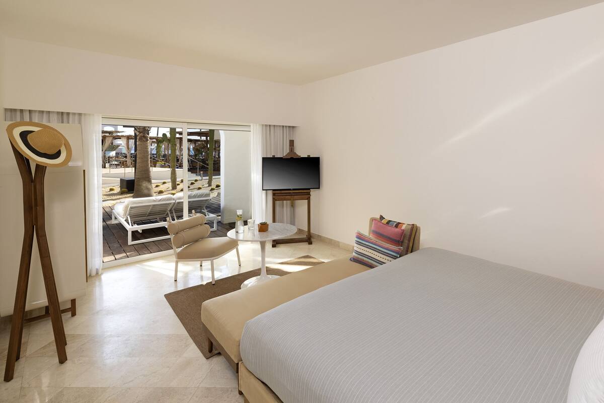 Quarto de hotel com cama, televisão, mesa pequena e vista para área externa com piscina e espreguiçadeiras.