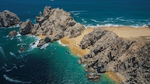 Vista aérea de um litoral rochoso com uma praia arenosa e águas azuis cristalinas.