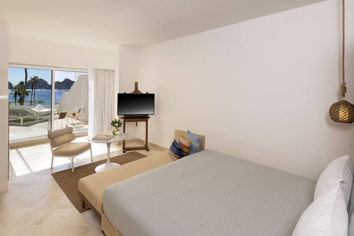 Quarto de hotel com cama, televisão, uma mesinha com uma planta, uma cadeira e vista para o mar.