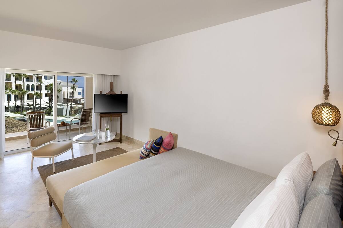 Quarto de hotel com cama, televisão, mesa com cadeiras e vista para palmeiras e edifícios lá fora.