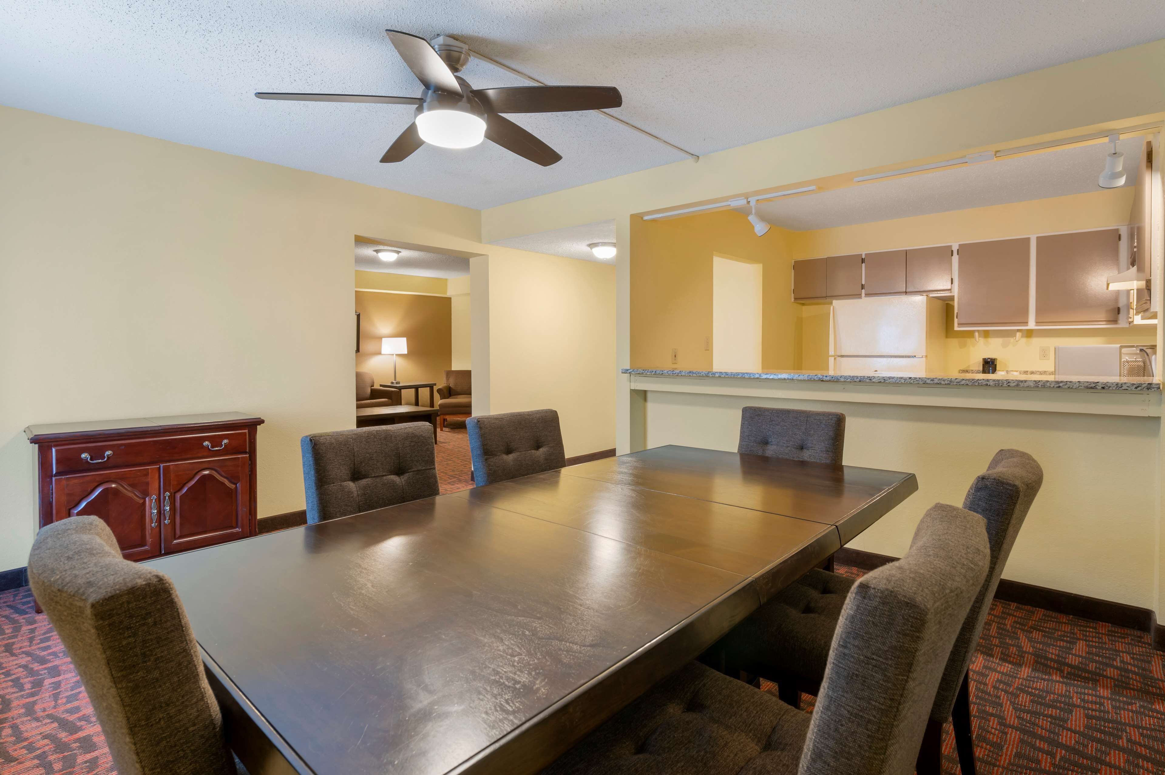 Sala de conferências com uma mesa grande, seis cadeiras, um ventilador de teto e uma kitchenette ao fundo.