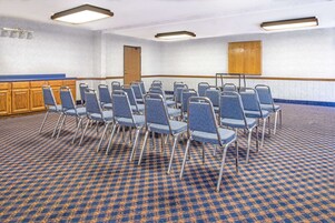 Uma sala de conferências com carpete estampado em azul, fileiras de cadeiras azuis, um balcão de madeira e uma porta.