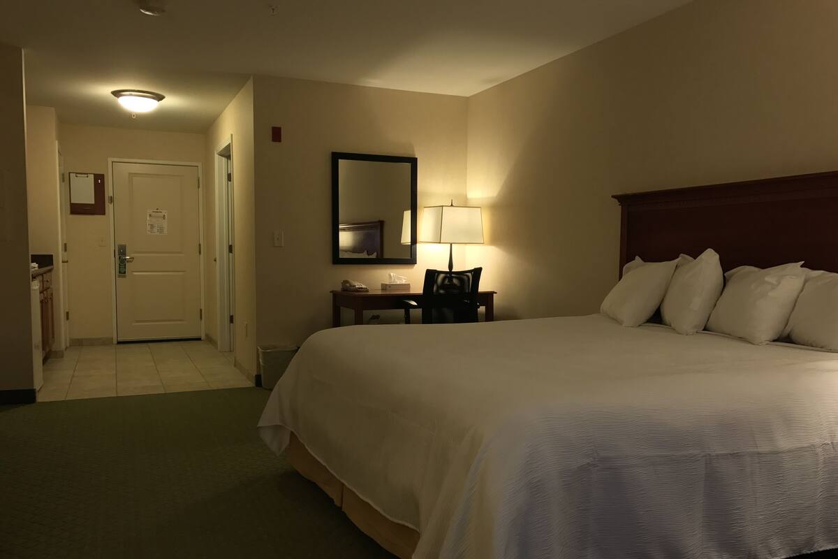 Quarto de hotel com uma cama grande, uma mesa de trabalho com luminária, uma cadeira e um espelho na parede.