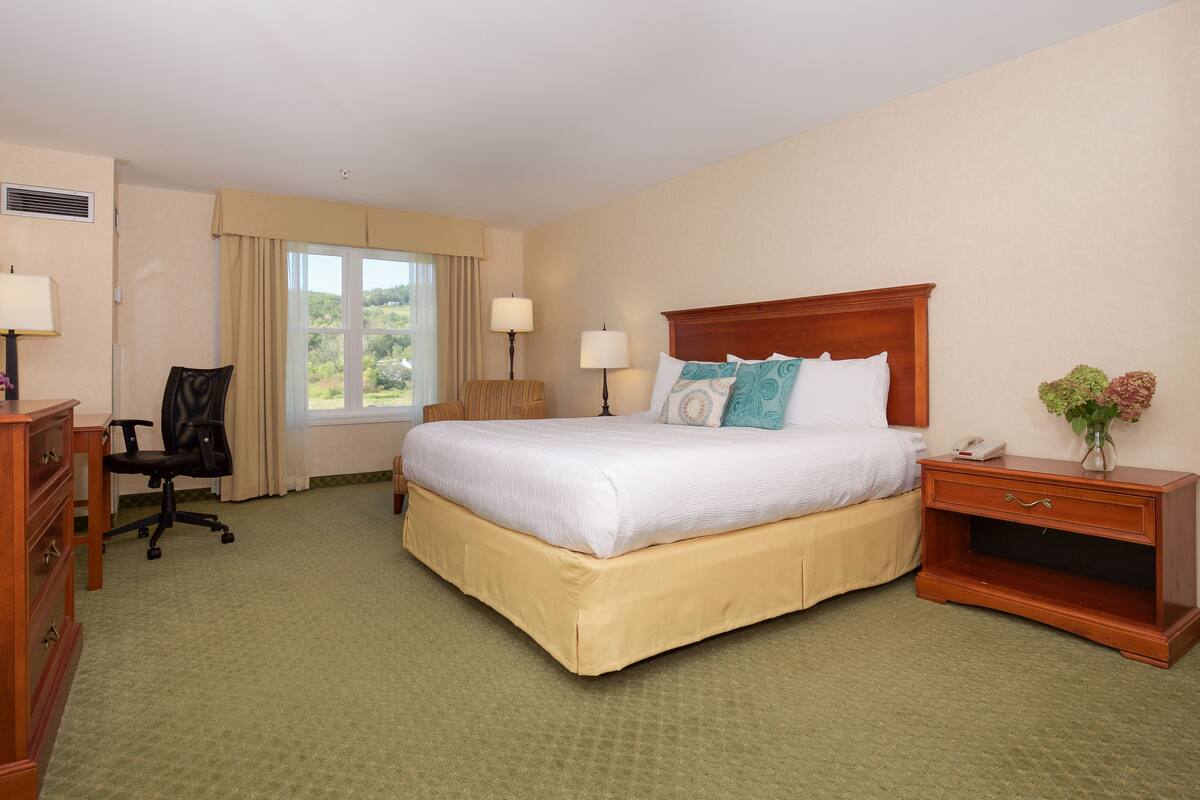 Quarto de hotel com uma cama grande, uma escrivaninha, uma cadeira, uma mesinha de cabeceira e uma janela com cortinas.