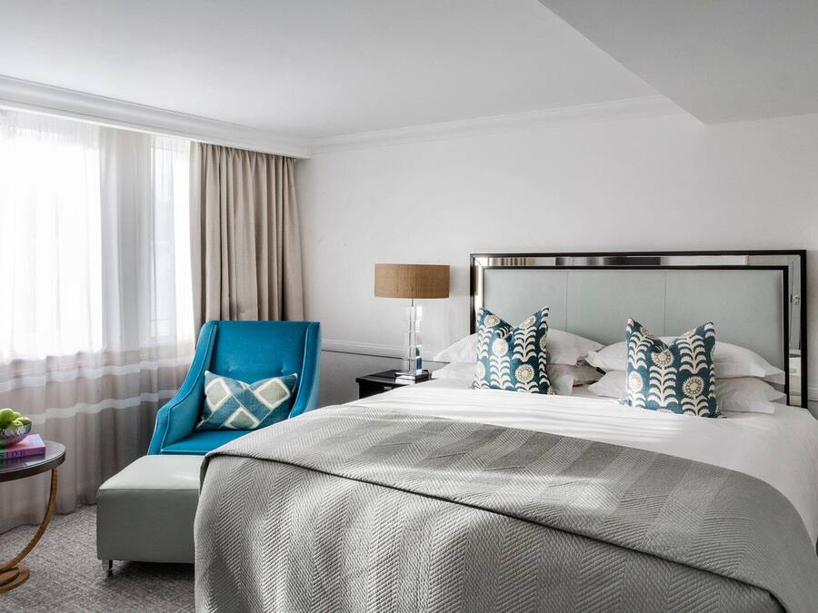 Quarto de hotel com uma cama grande, uma poltrona azul, uma pequena mesa de apoio com livros e uma mesinha de cabeceira com abajur.