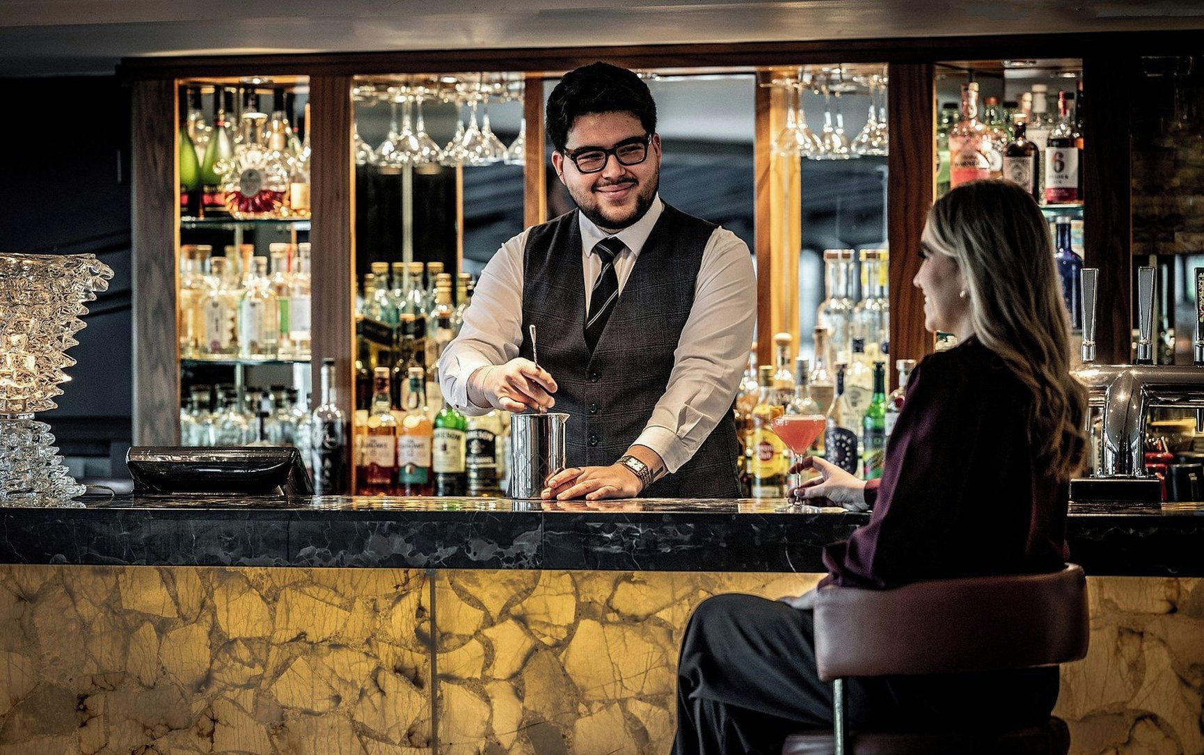 Ambiente de bar com um barman preparando uma bebida, um cliente sentado e um balcão de bar bem abastecido.