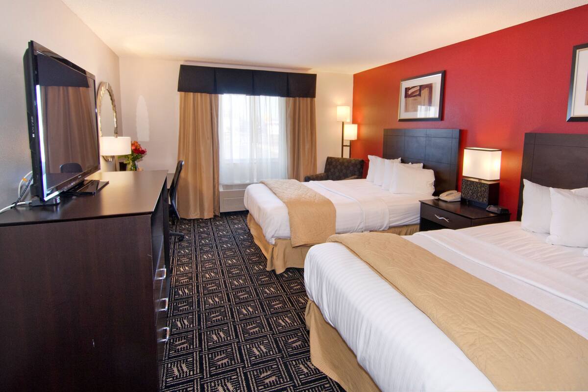 Quarto de hotel com duas camas, uma mesa de escritório com televisão, uma cadeira, um abajur e uma janela com cortinas.
