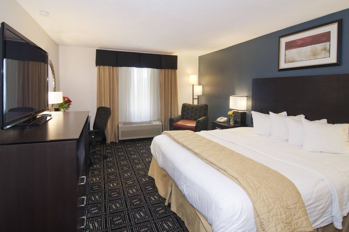 Quarto de hotel com uma cama grande, uma escrivaninha com televisão, uma cadeira, uma luminária de mesa e uma janela com cortinas.