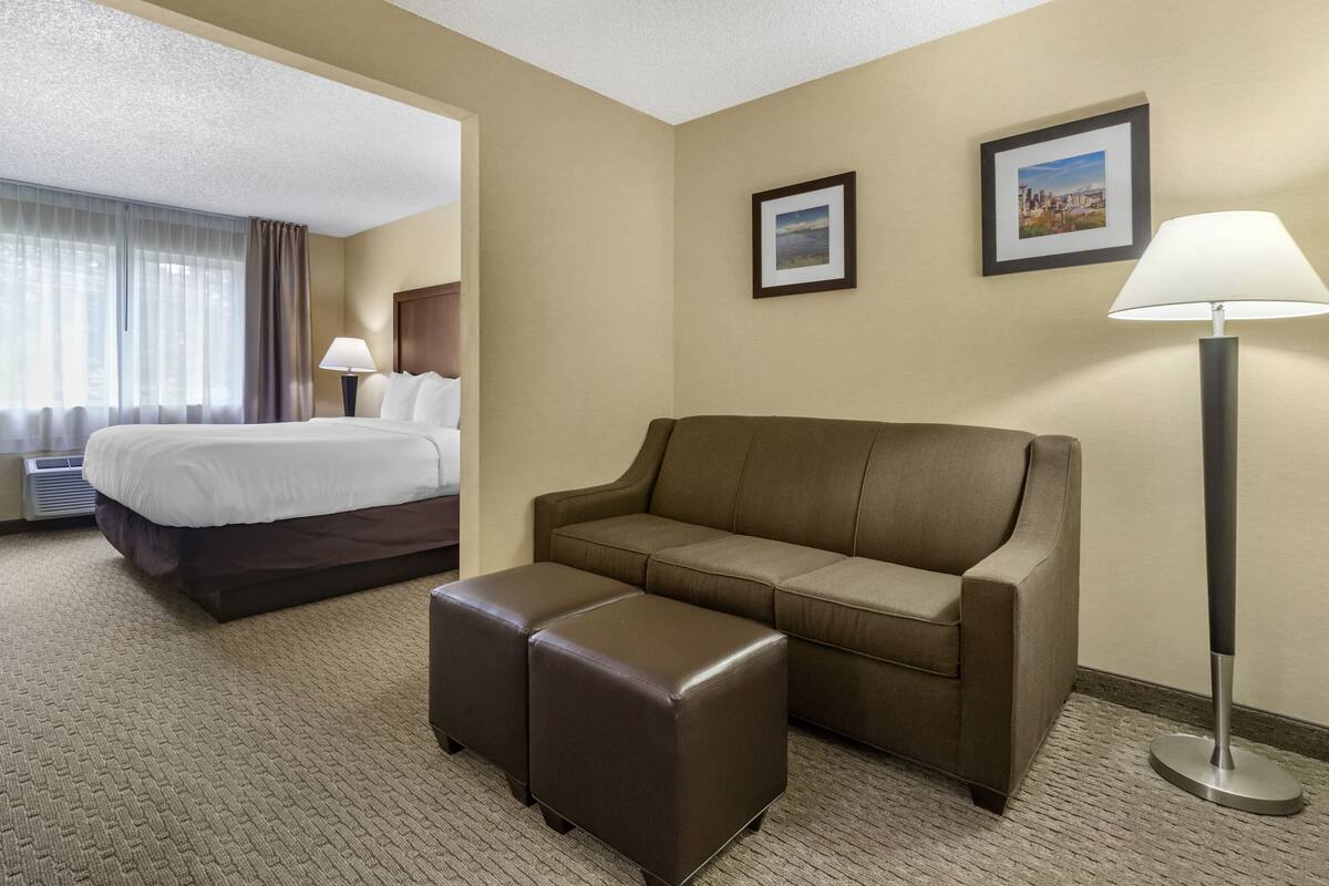 Quarto de hotel com cama, sofá, banquinho e duas luminárias.
