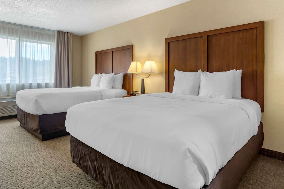 Quarto de hotel com duas camas, cabeceira de madeira, mesa de cabeceira com luminária e uma janela grande com cortinas.