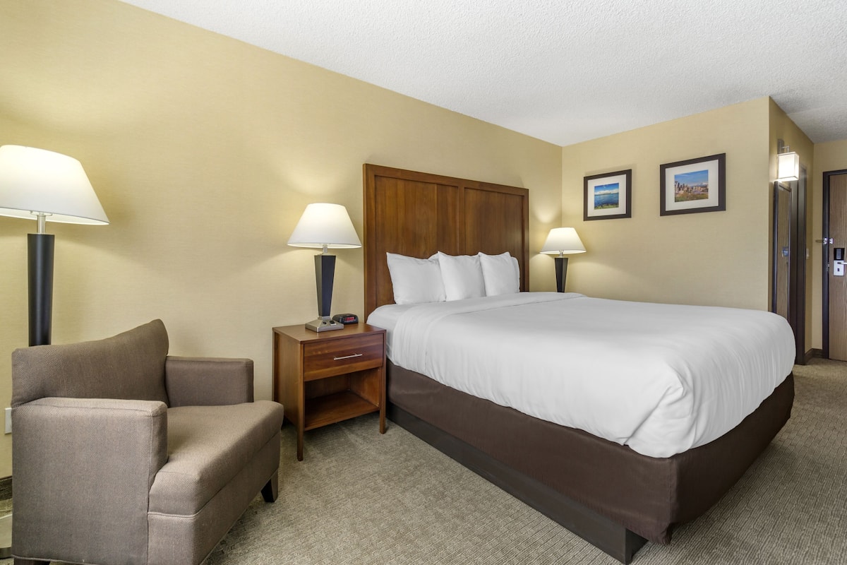 Quarto de hotel com uma cama grande, uma cadeira, duas luminárias de cabeceira, uma mesinha de cabeceira e duas fotos emolduradas na parede.