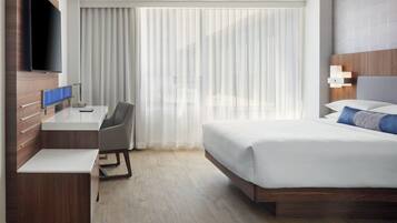Quarto de hotel com cama, escrivaninha, cadeira, televisão e janela com persianas.