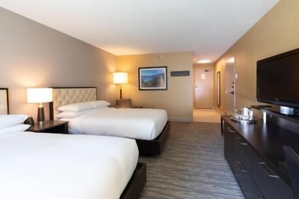 Quarto de hotel com duas camas, televisão, escrivaninha com luminária e um quadro na parede.