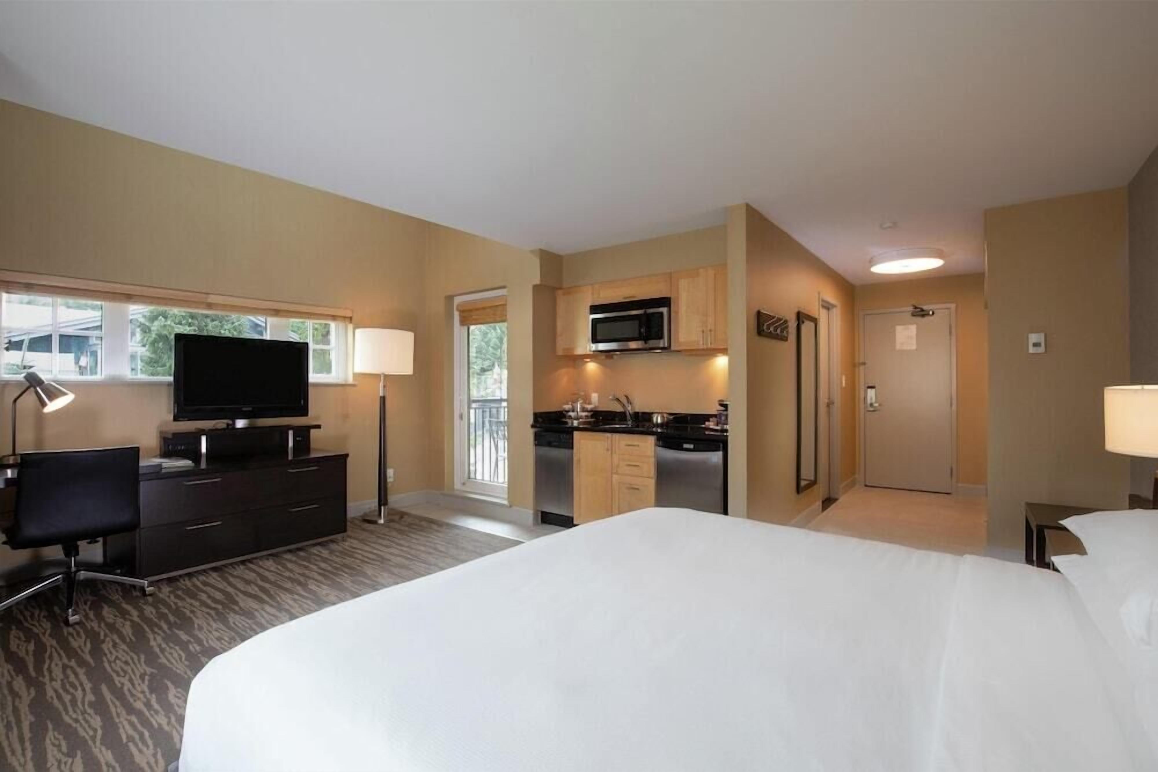 Quarto de hotel com cama, televisão, escrivaninha, cadeira, cozinha compacta e janela.