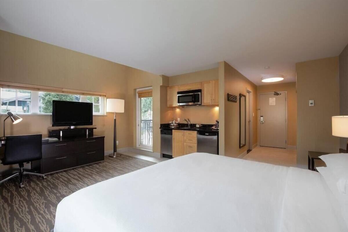 Quarto de hotel com cama, televisão, escrivaninha, cadeira, cozinha compacta e janela.