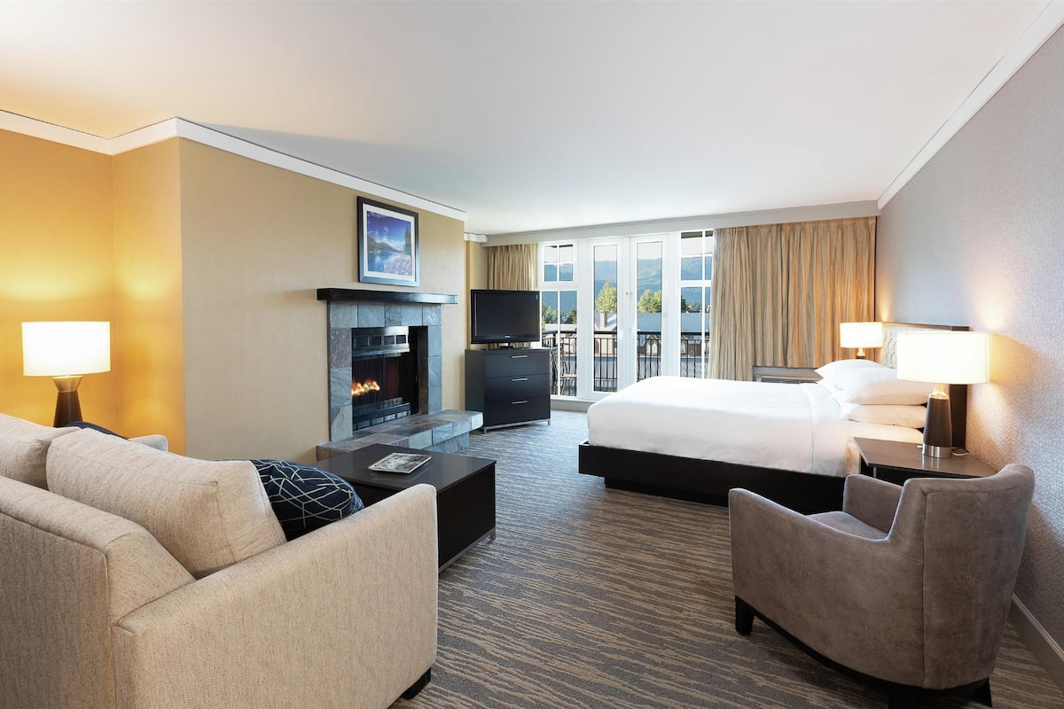 Quarto de hotel com cama, sofá, televisão, lareira e varanda com cortinas.