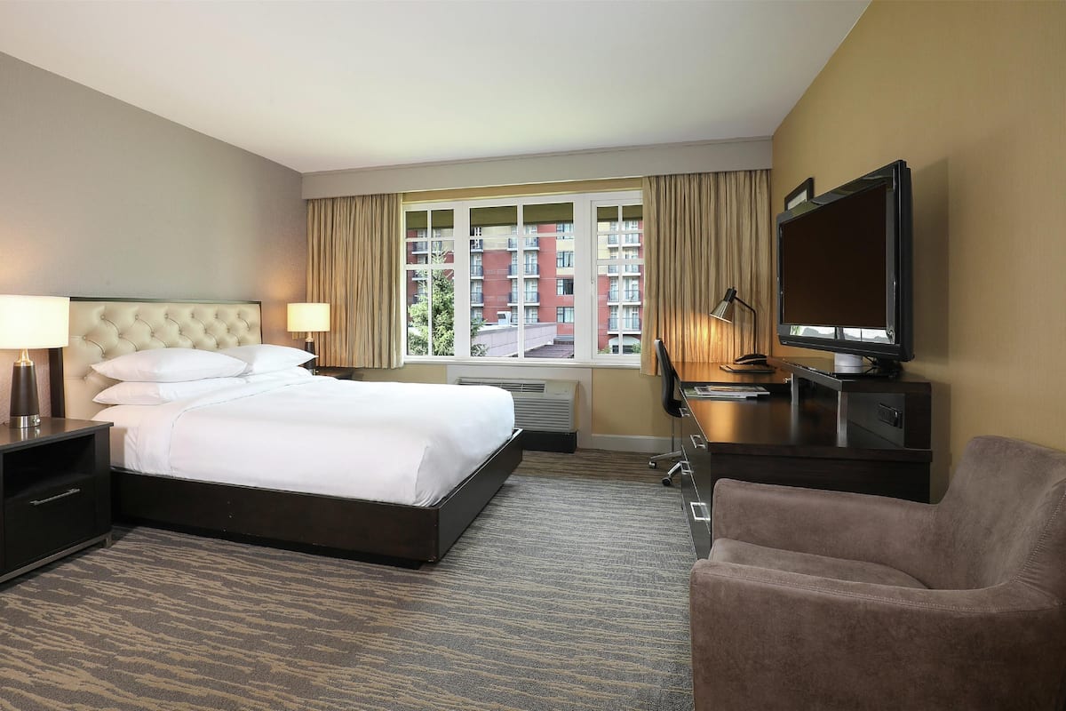 Quarto de hotel com uma cama, duas luminárias de cabeceira, uma escrivaninha com uma televisão, uma cadeira e vista para prédios pela janela.