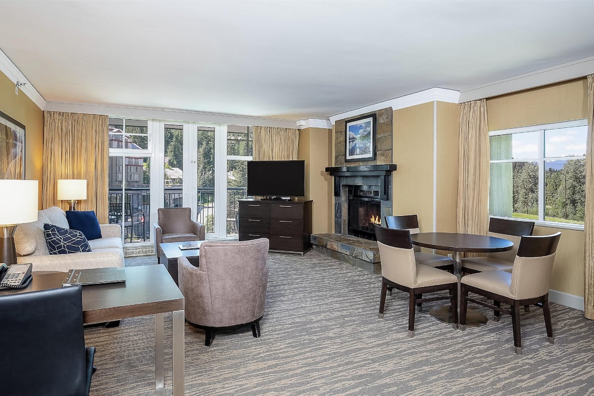 Quarto de hotel com sofá, poltronas, mesa de jantar, televisão, lareira e grandes janelas com vista para o exterior.
