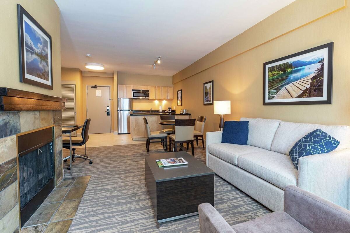 Quarto de hotel com sofá, mesa de centro, lareira, cozinha e mesa de jantar.