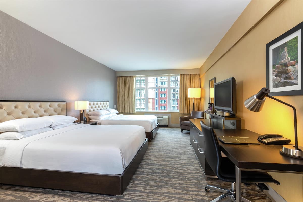 Quarto de hotel com duas camas, uma escrivaninha, uma cadeira, televisão e janela com vista para edifícios.