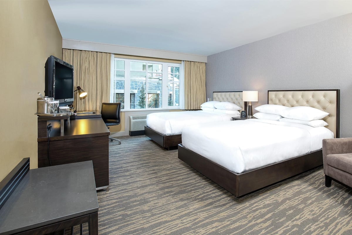 Quarto de hotel com duas camas, uma escrivaninha, uma cadeira, uma televisão e uma janela com vista.