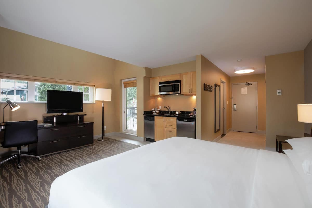 Quarto de hotel com cama, escrivaninha com televisão, cozinha compacta e janela com vista.