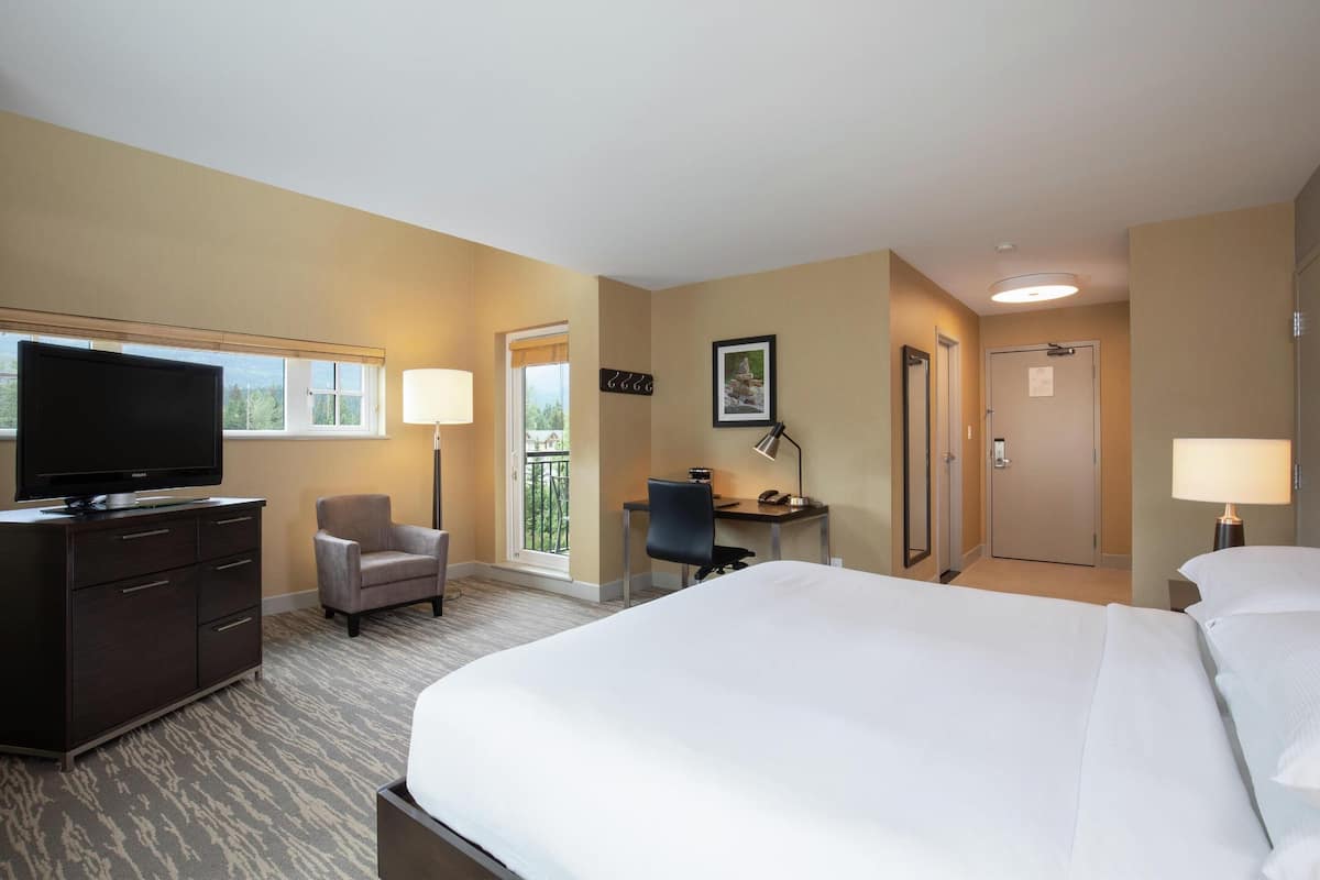 Quarto de hotel com uma cama grande, uma televisão num aparador, uma mesa de trabalho com uma cadeira e uma porta para uma varanda.