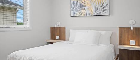 Quarto com cama, roupa de cama branca, obra de arte na parede, luminárias de cabeceira e janela com persianas.