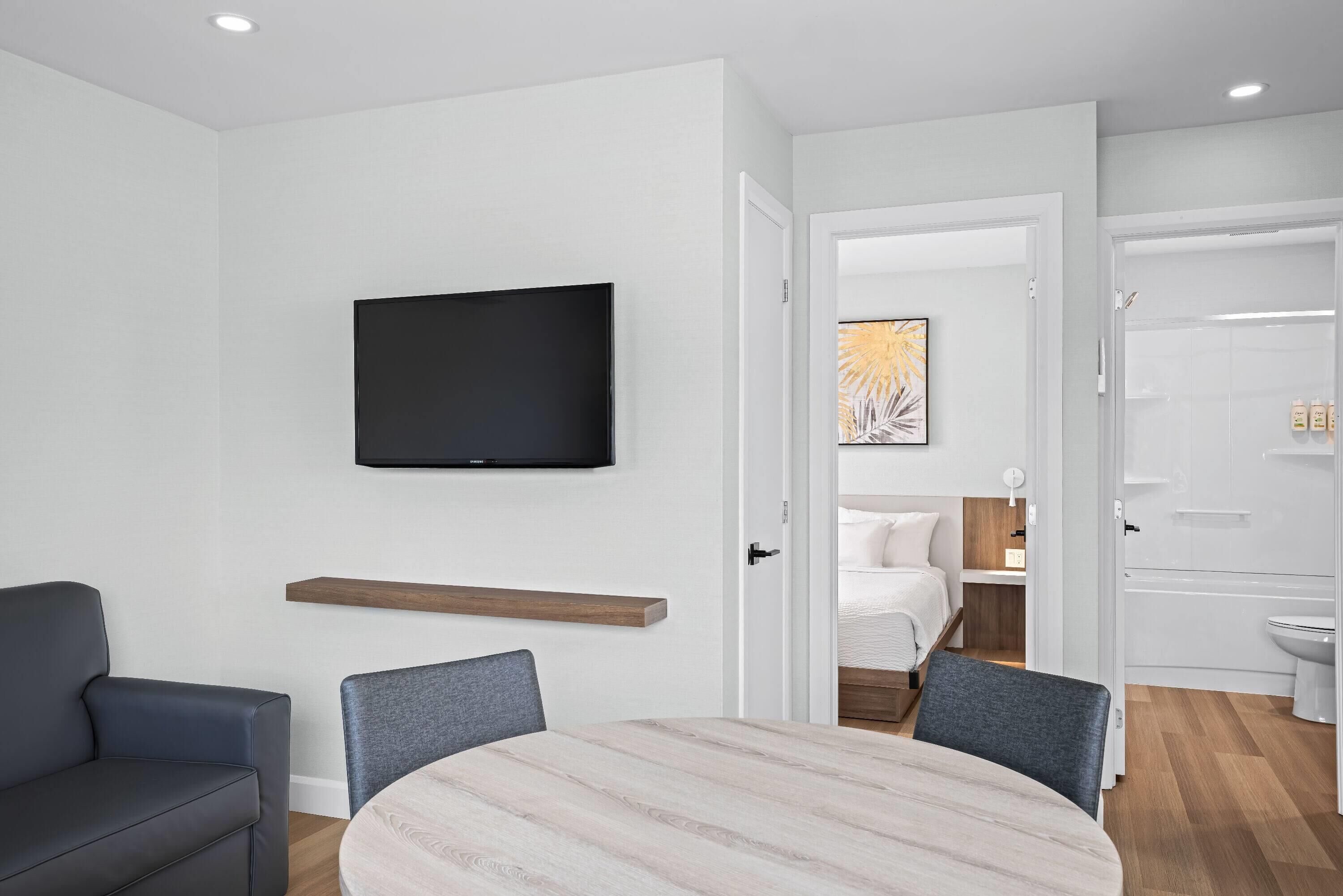 Quarto de hotel moderno com televisão de tela plana na parede, prateleira de madeira, mesa de jantar redonda com cadeiras, cama com roupa de cama branca e banheiro com vaso sanitário e chuveiro.