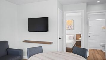 Quarto de hotel moderno com televisão de tela plana na parede, prateleira de madeira, mesa de jantar redonda com cadeiras, cama com roupa de cama branca e banheiro com vaso sanitário e chuveiro.