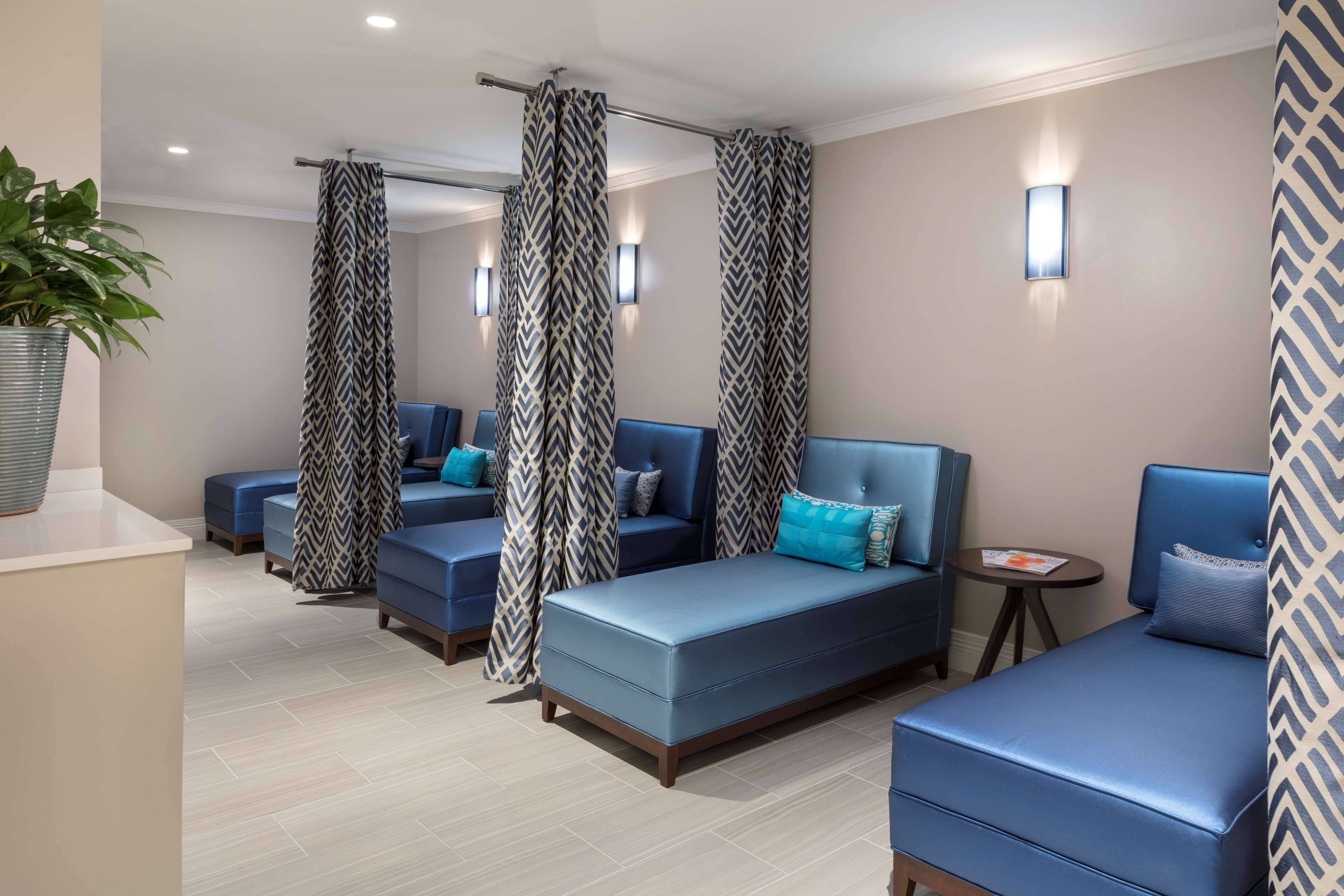 Quarto de hotel moderno com uma cama grande, dois sofás azuis, cortinas estampadas e uma pequena mesa com uma planta.