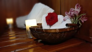 Um tapete de banho de madeira, uma vela, um cesto com uma rosa vermelha, toalhas brancas e flores cor-de-rosa.