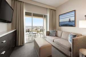 Quarto de hotel com sofá, televisão, banco, mesa de jantar, cadeiras e uma grande janela com vista para o oceano.
