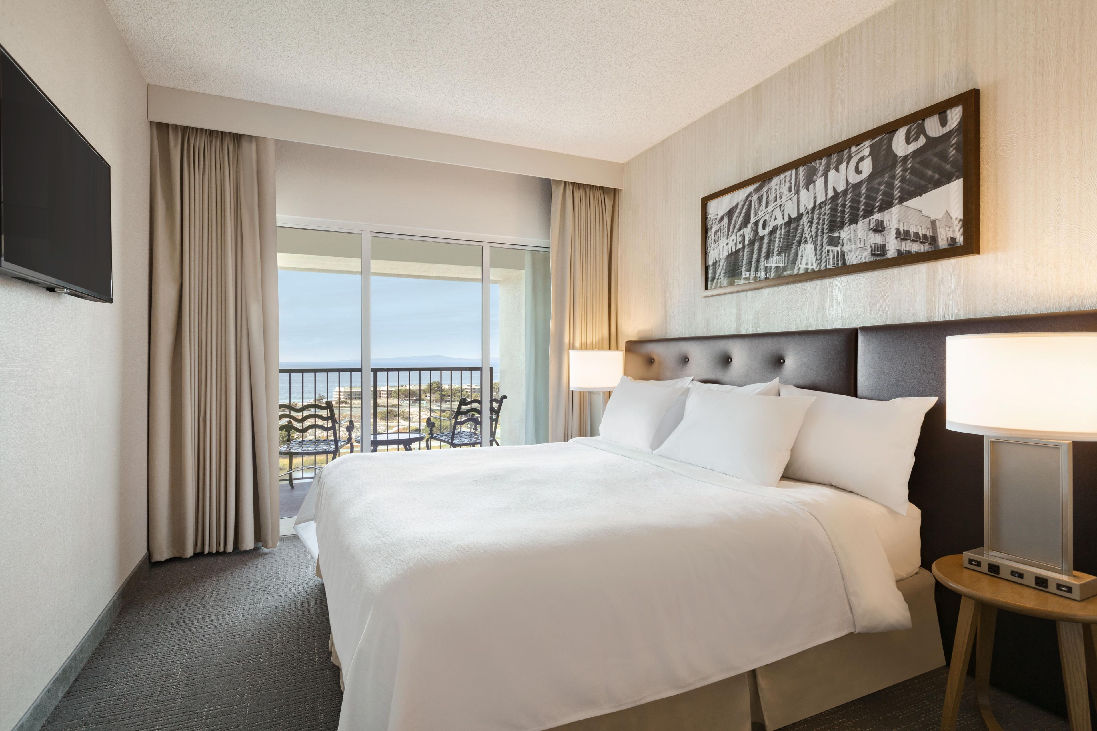 Quarto de hotel com cama, duas luminárias, televisão, janela com cortinas e um quadro na parede.