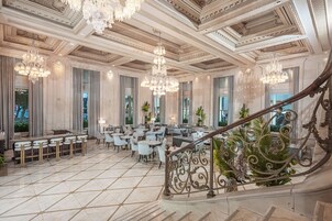 Um hall imponente com piso de mármore, lustres elegantes e uma escadaria com grades ornamentadas.