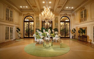 Um hall luxuoso com um imponente candelabro, amplas janelas em arco e um arranjo floral central sobre uma mesa espelhada.