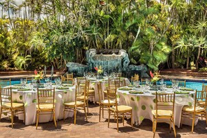 Área de refeições ao ar livre com mesas preparadas para uma refeição, rodeada por vegetação tropical exuberante e uma piscina.