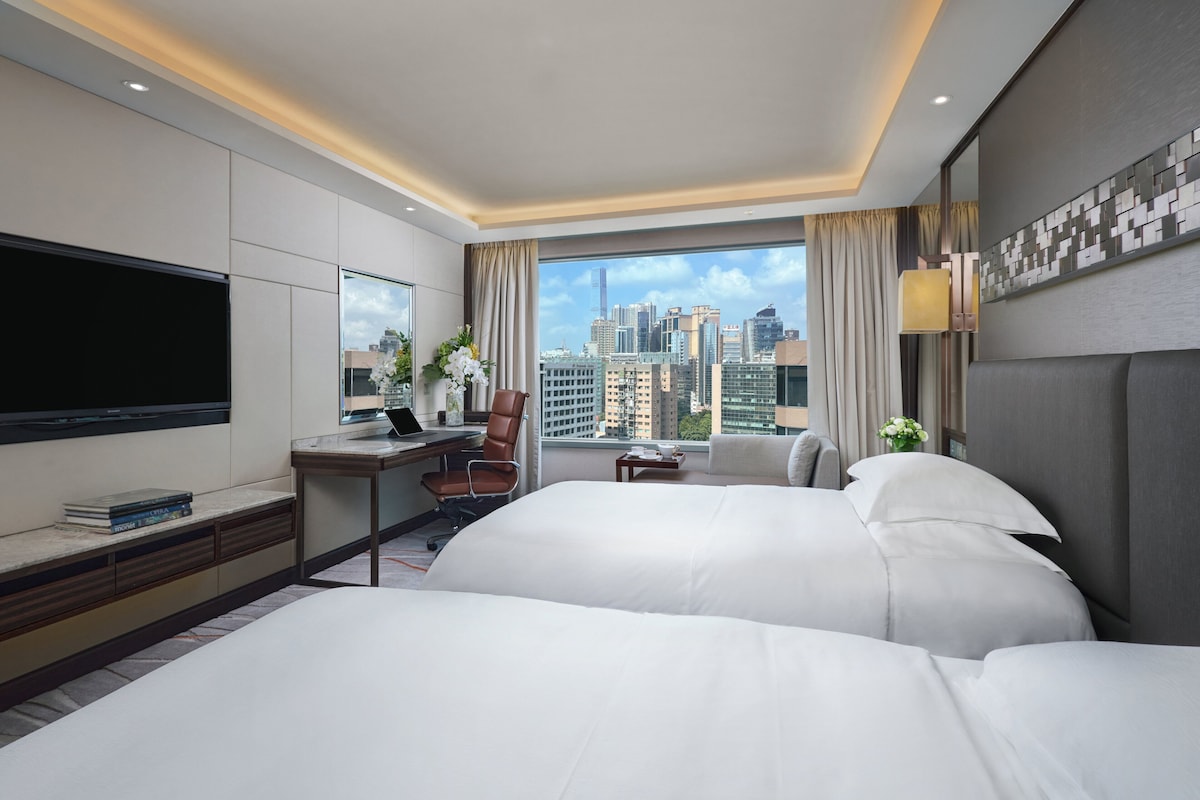 Quarto de hotel com uma cama grande, uma mesa com cadeira, uma televisão e vista para a cidade.