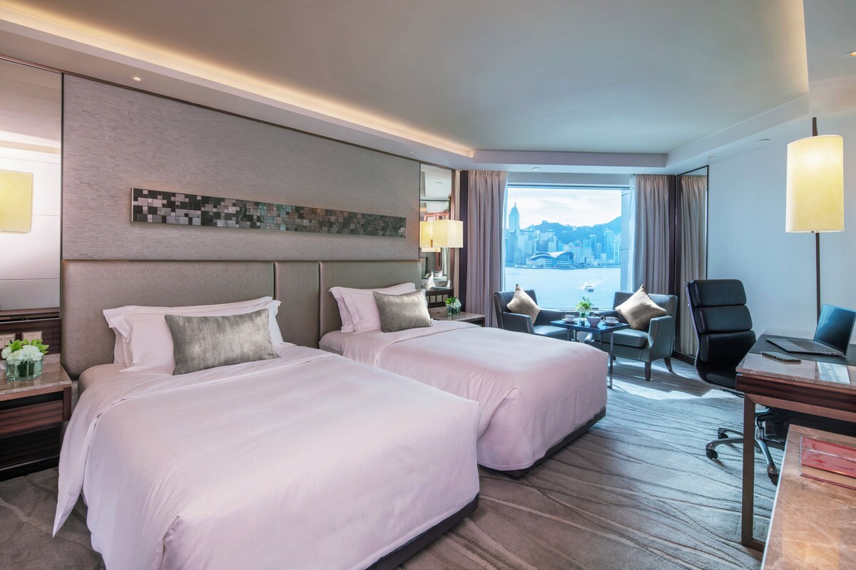 Quarto de hotel com duas camas, uma escrivaninha, uma cadeira e vista para a paisagem urbana.